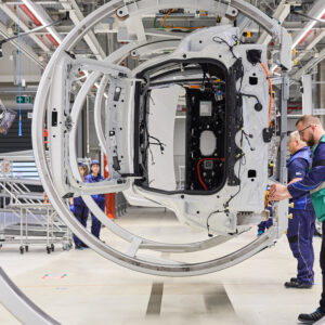 Workers look at a BMW chassis on a rotating jig at a factory.